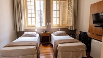 Quarto de hotel com duas camas de solteiro, uma pequena mesa de cabeceira com abajur, televisão e mini-geladeira.