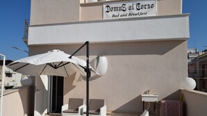 Uma varanda com móveis de vime branco, um guarda-sol branco e uma placa indicando "Domus al Corso Bed and Breakfast".