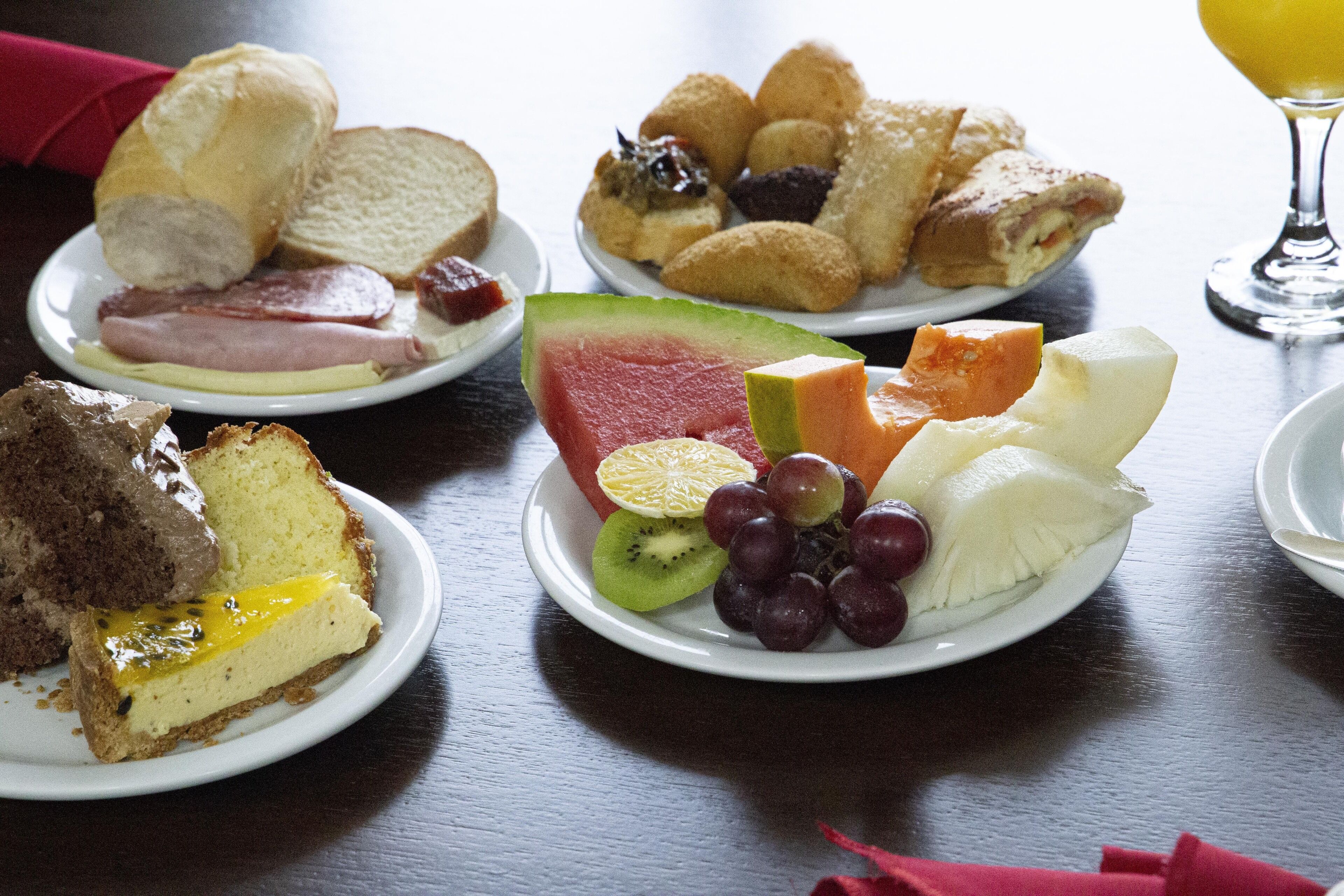 Três pratos de comida com pães, presunto, queijo e frutas diversas.