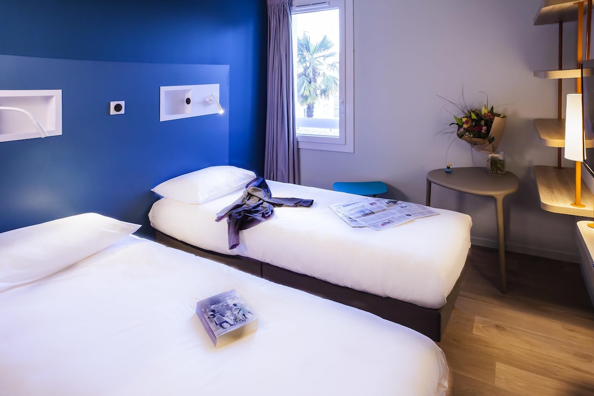 Quarto de hotel com duas camas, uma mesa pequena, uma luminária e uma janela com vista para palmeiras.
