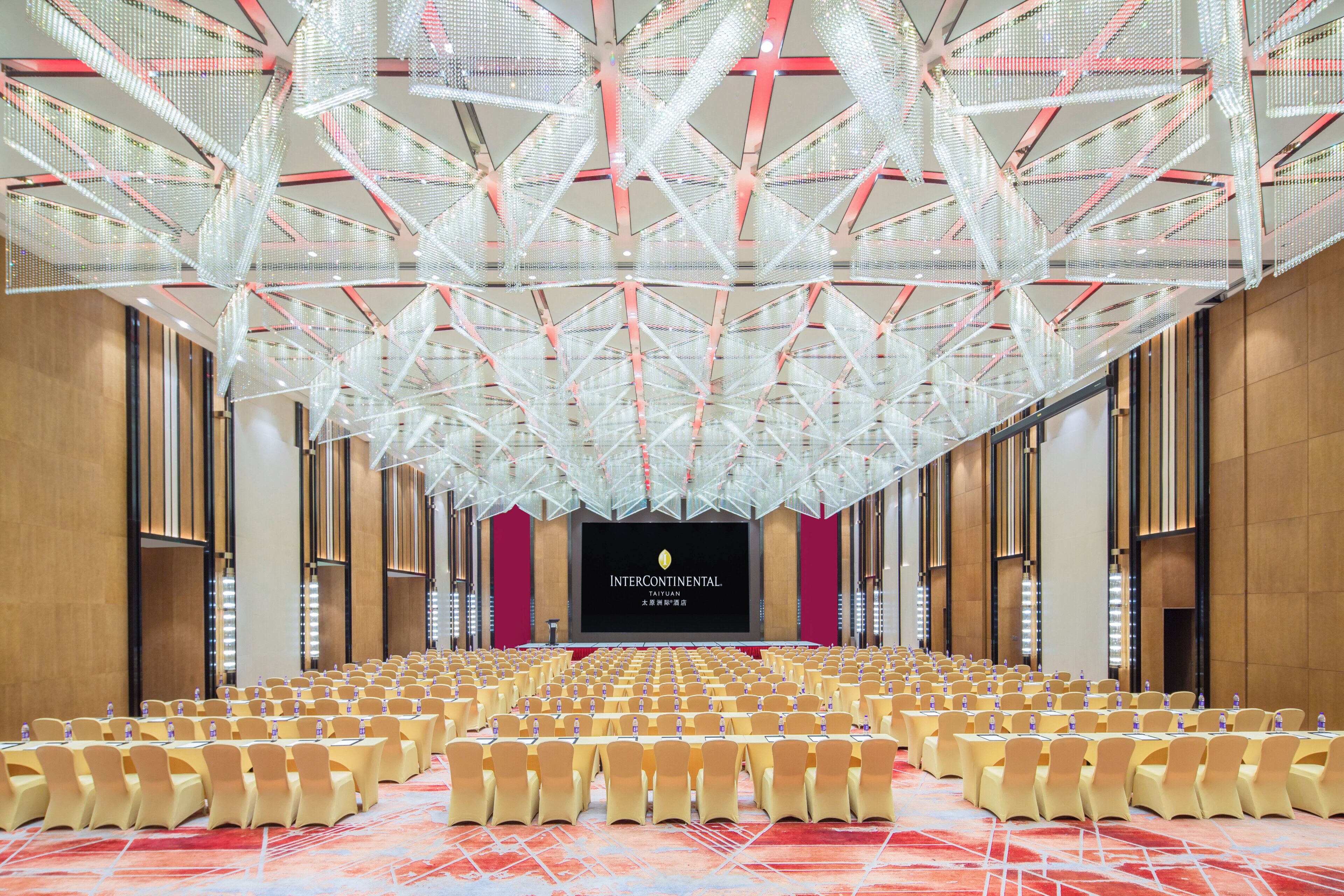 Uma sala de conferências com fileiras de cadeiras, um palco com uma tela e um teto com luminária geométrica.