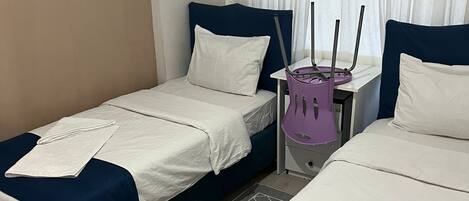 Quarto de hotel com duas camas, uma mesinha e uma cadeira roxa, além de uma janela com cortinas brancas.