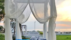 Cama com colchão azul e dossel branco, travesseiros e velas sobre um deck de madeira, com vista para o mar e edifícios.