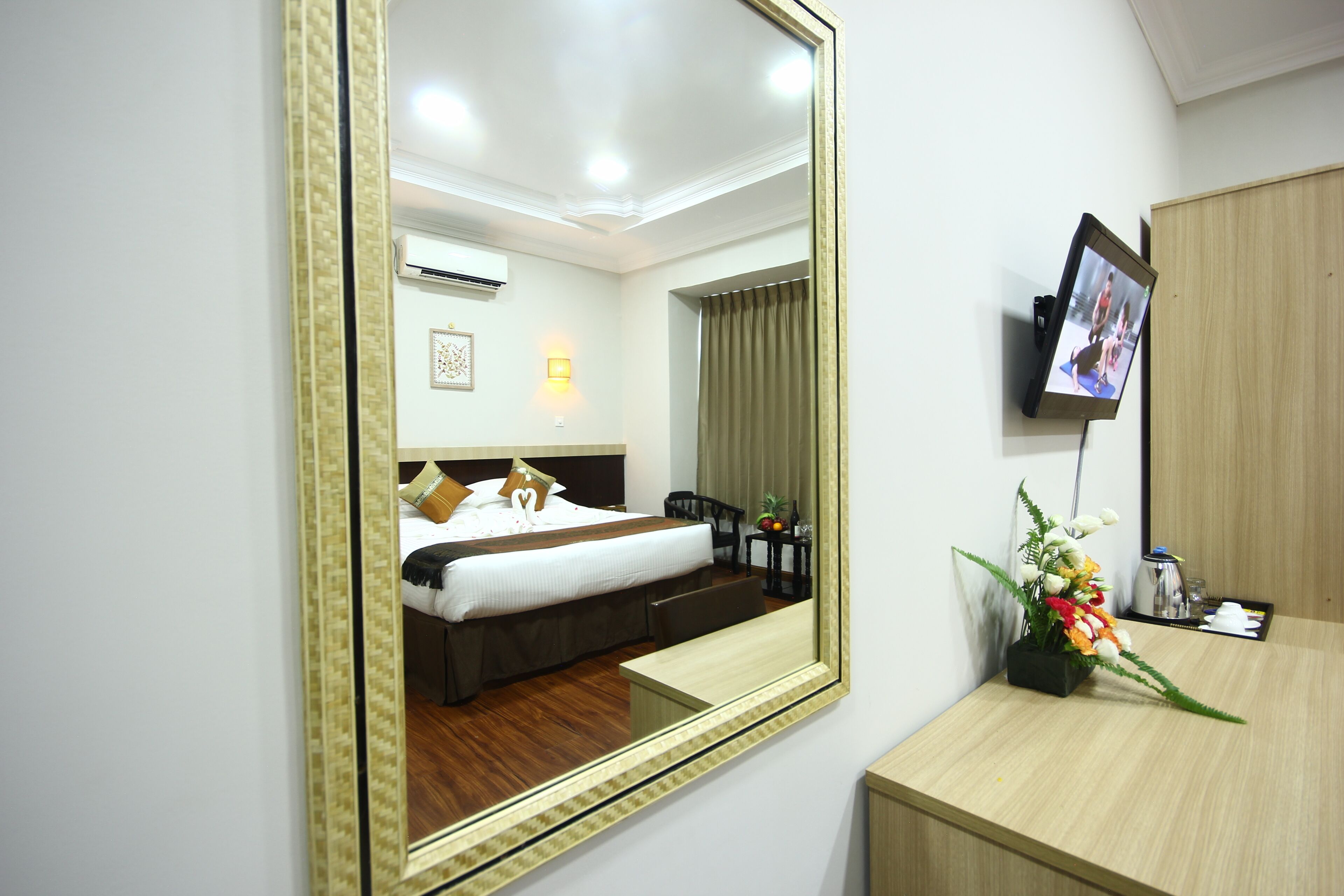 Quarto de hotel com um espelho grande, cama com lençóis brancos, televisão fixada na parede e uma pequena mesa com um vaso de flores.