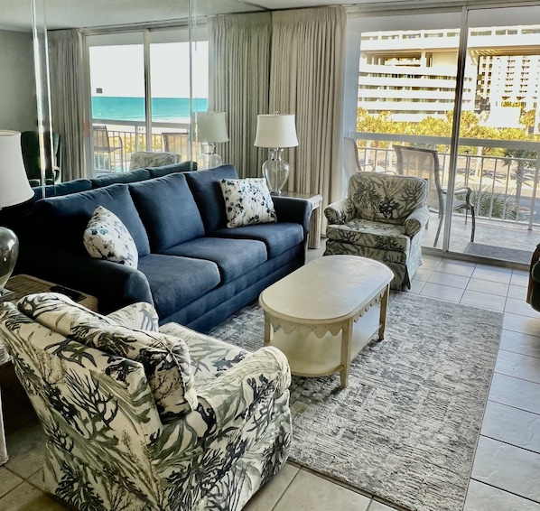 Uma sala de estar moderna com sofá, poltrona e mesa de centro, janelas amplas com vista para o oceano e um prédio, e uma varanda com guarda-corpo.