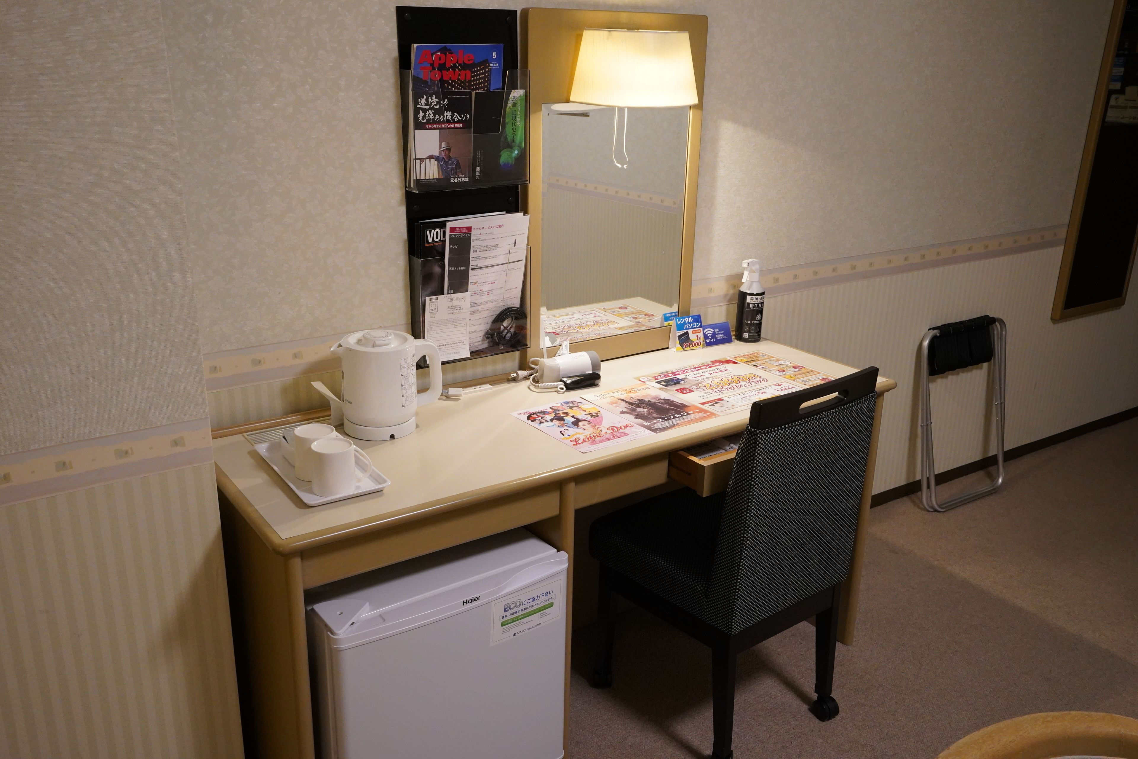 Quarto de hotel com escrivaninha, cadeira, espelho, luminária e pequena geladeira.