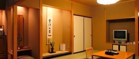 Um quarto tradicional em estilo japonês, com tatames, uma mesa baixa com almofadas, televisão e portas deslizantes.