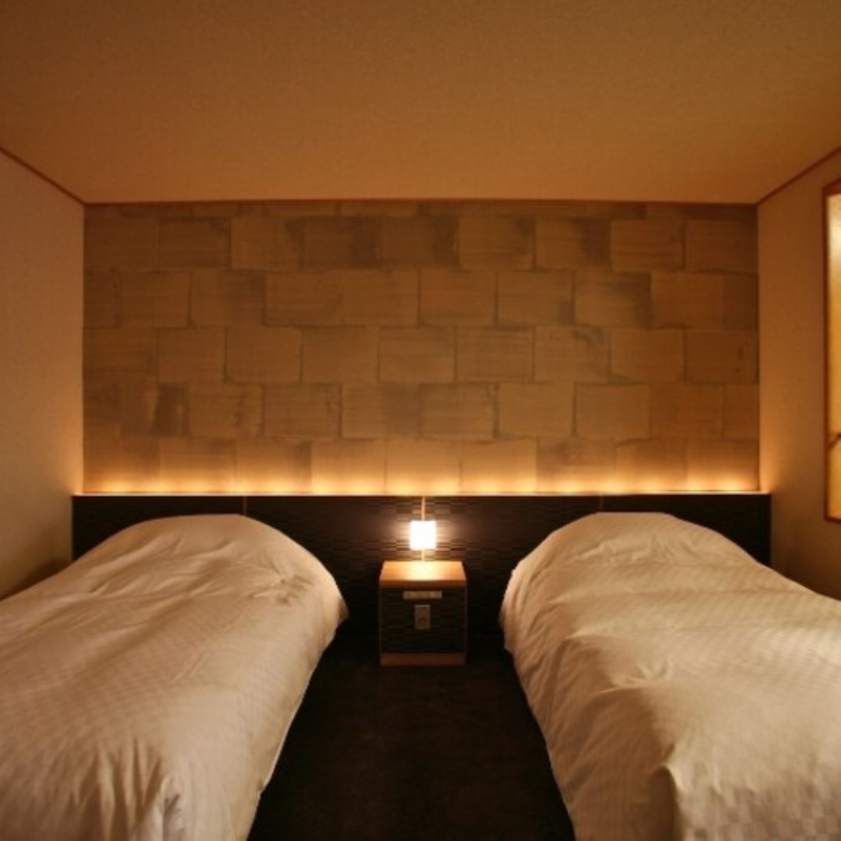 Quarto de hotel com duas camas de solteiro, uma mesa de cabeceira e parede com padrão de tijolos.