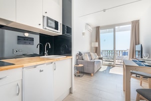 Cozinha moderna com micro-ondas, pia e armários brancos. Há uma área de refeições com mesa e cadeiras, e uma varanda com vista para o mar.