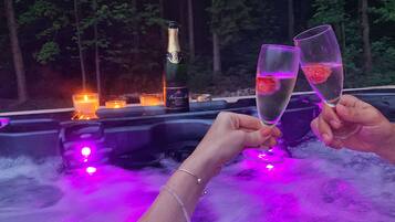 Uma pessoa segurando duas taças de champanhe com uma garrafa de champanhe em uma superfície aquecida, rodeada por velas e um cenário florestal.