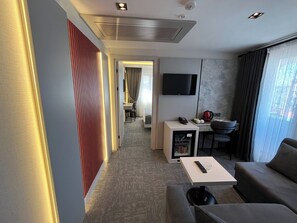 Quarto de hotel com sofá, uma pequena mesa, televisão, cozinha compacta e banheiro ao fundo.