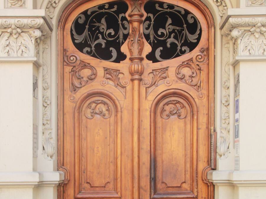 Uma porta de madeira grande e ricamente decorada, com esculturas intrincadas, ladeada por colunas ornamentais e um arco detalhado.
