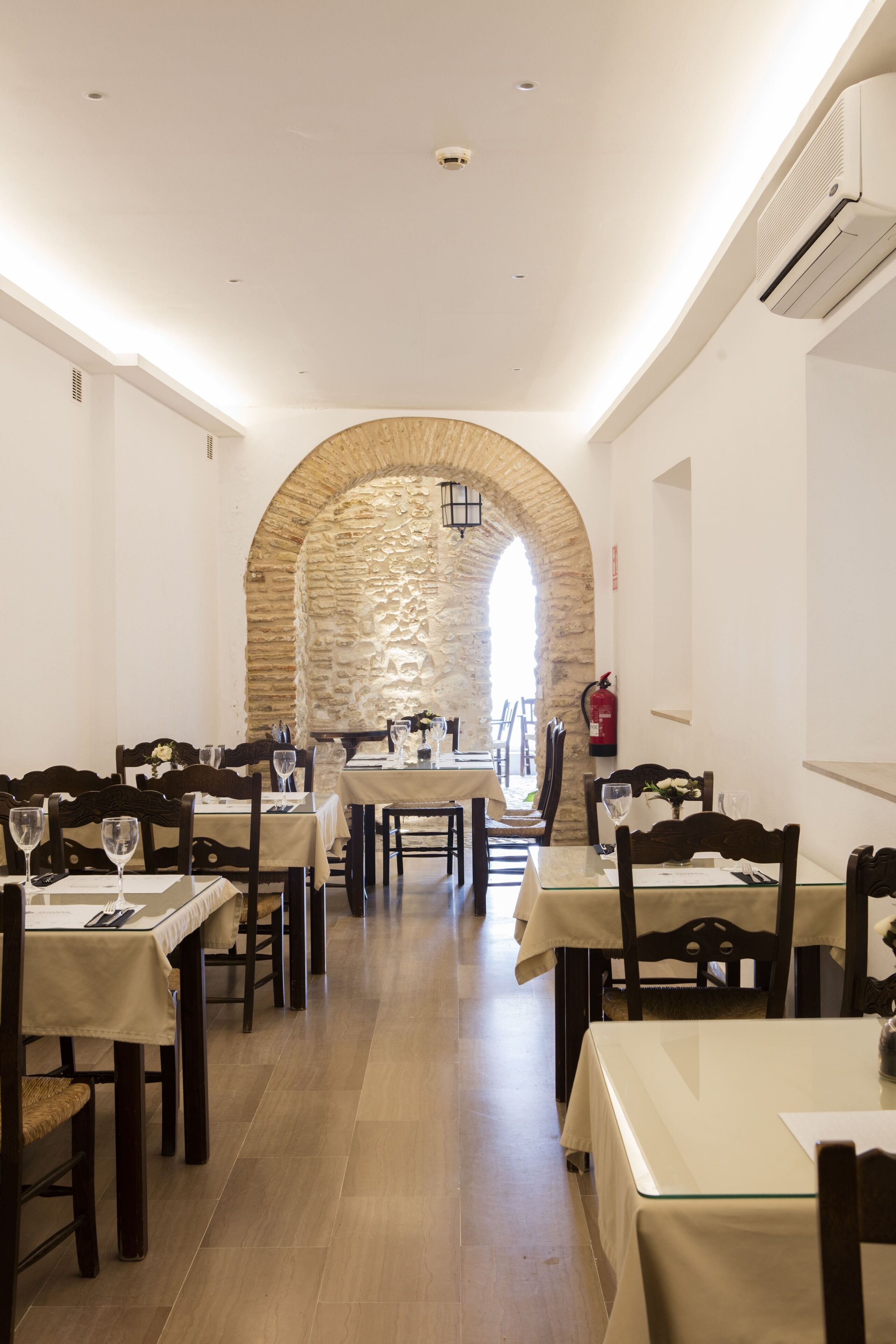 Um restaurante com várias mesas e cadeiras, um arco de pedra e um extintor de incêndio na parede.