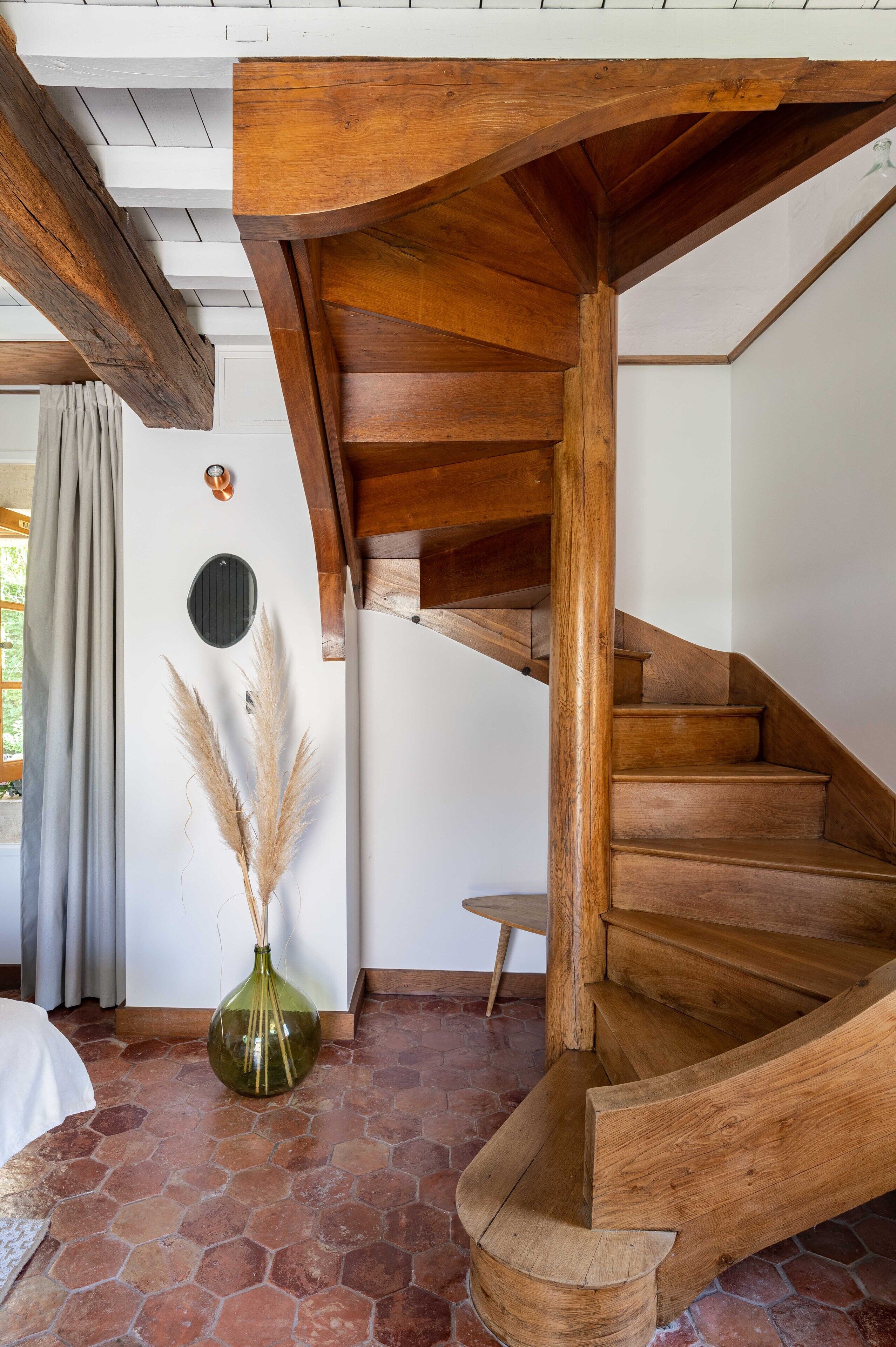 Escada em espiral de madeira com design rústico, um vaso com plumas de capim-do-campo e piso de azulejos hexagonais.