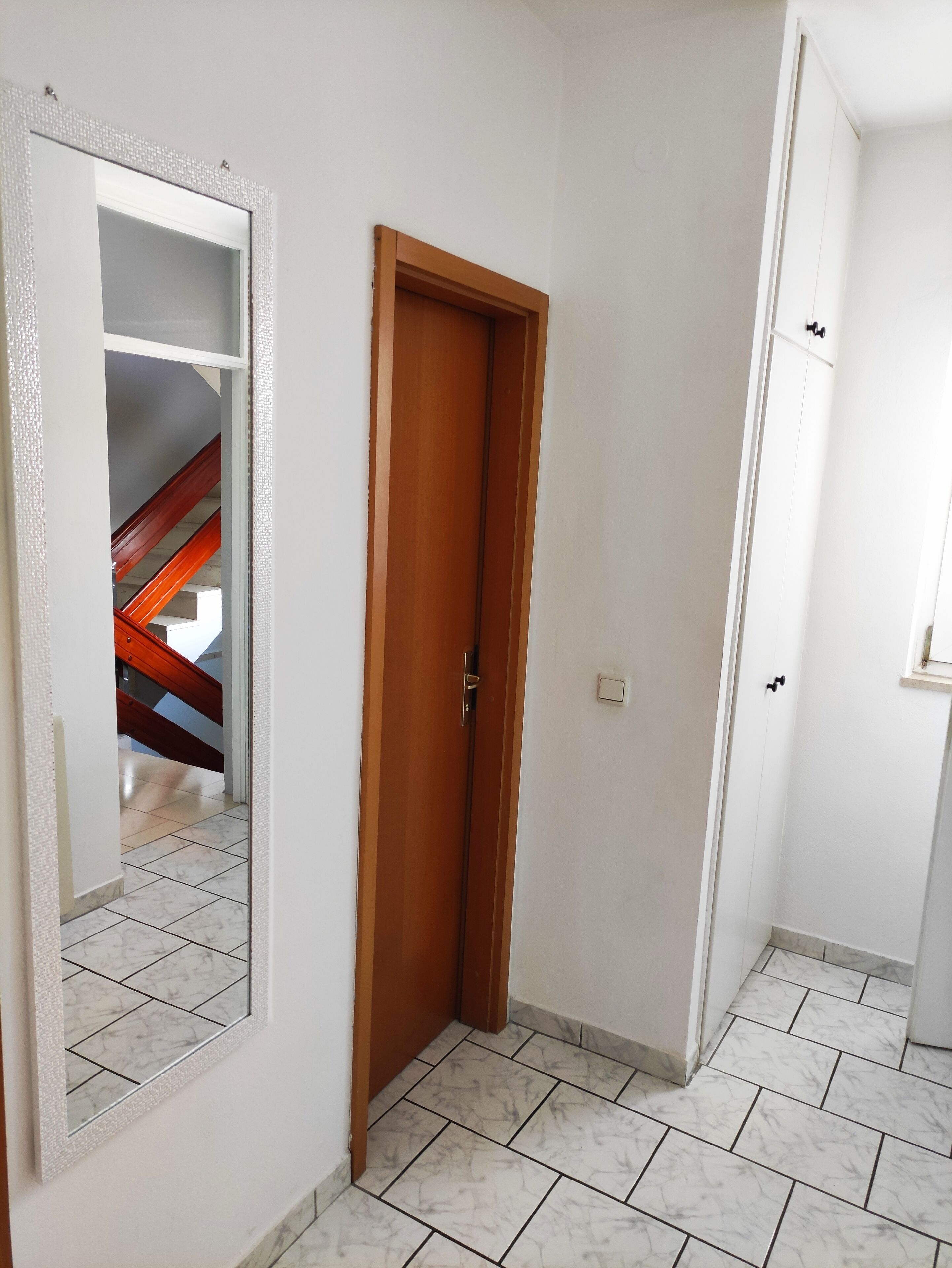 Um corredor com porta de madeira, parede branca e chão de azulejos.