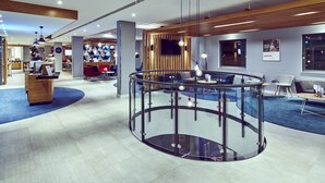 Um hall de hotel moderno com guarda-corpo de vidro central, áreas de estar e um balcão de recepção.