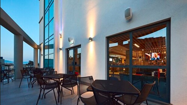 Uma área de refeições ao ar livre com mesas e cadeiras, grandes janelas e vista para o restaurante.