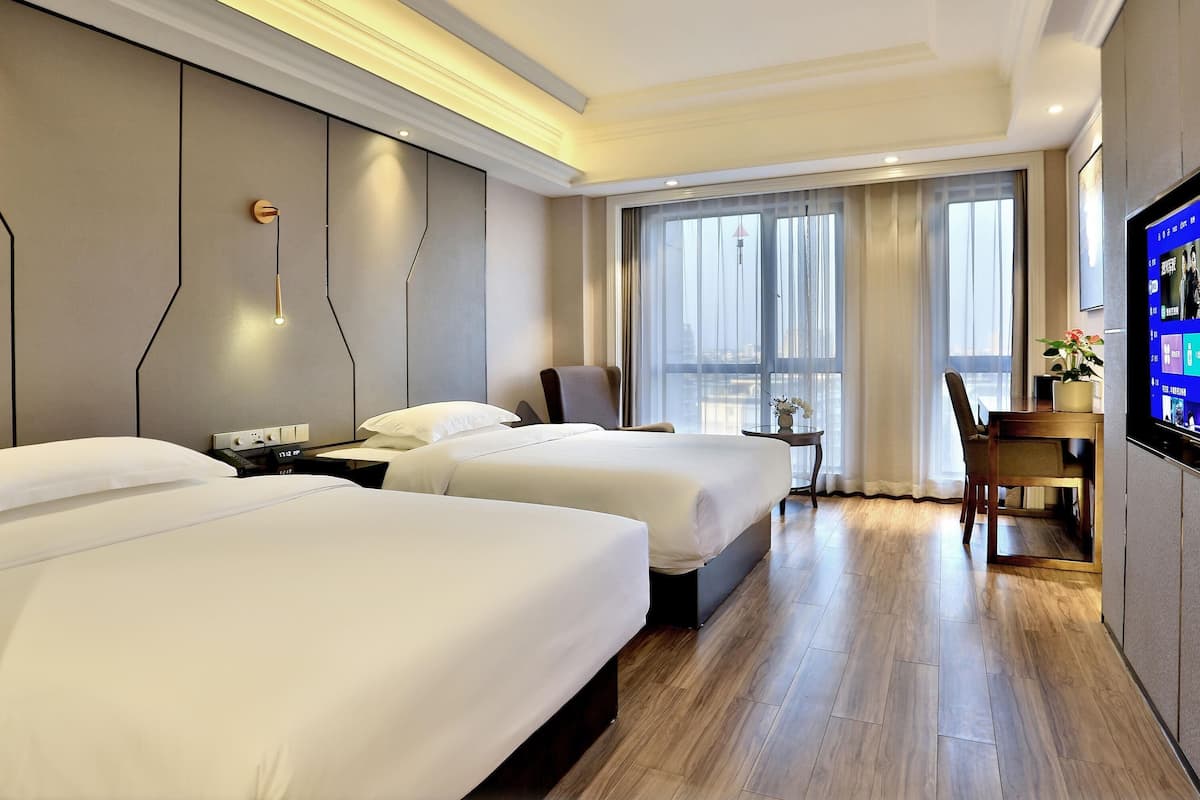 Quarto de hotel com duas camas, uma escrivaninha, uma cadeira, televisão e uma janela grande com cortinas.