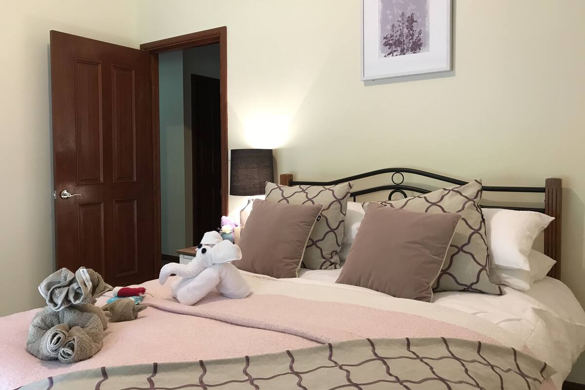 Cama bem arrumada com almofadas estampadas e manta rosa, porta de madeira e um quadro na parede.