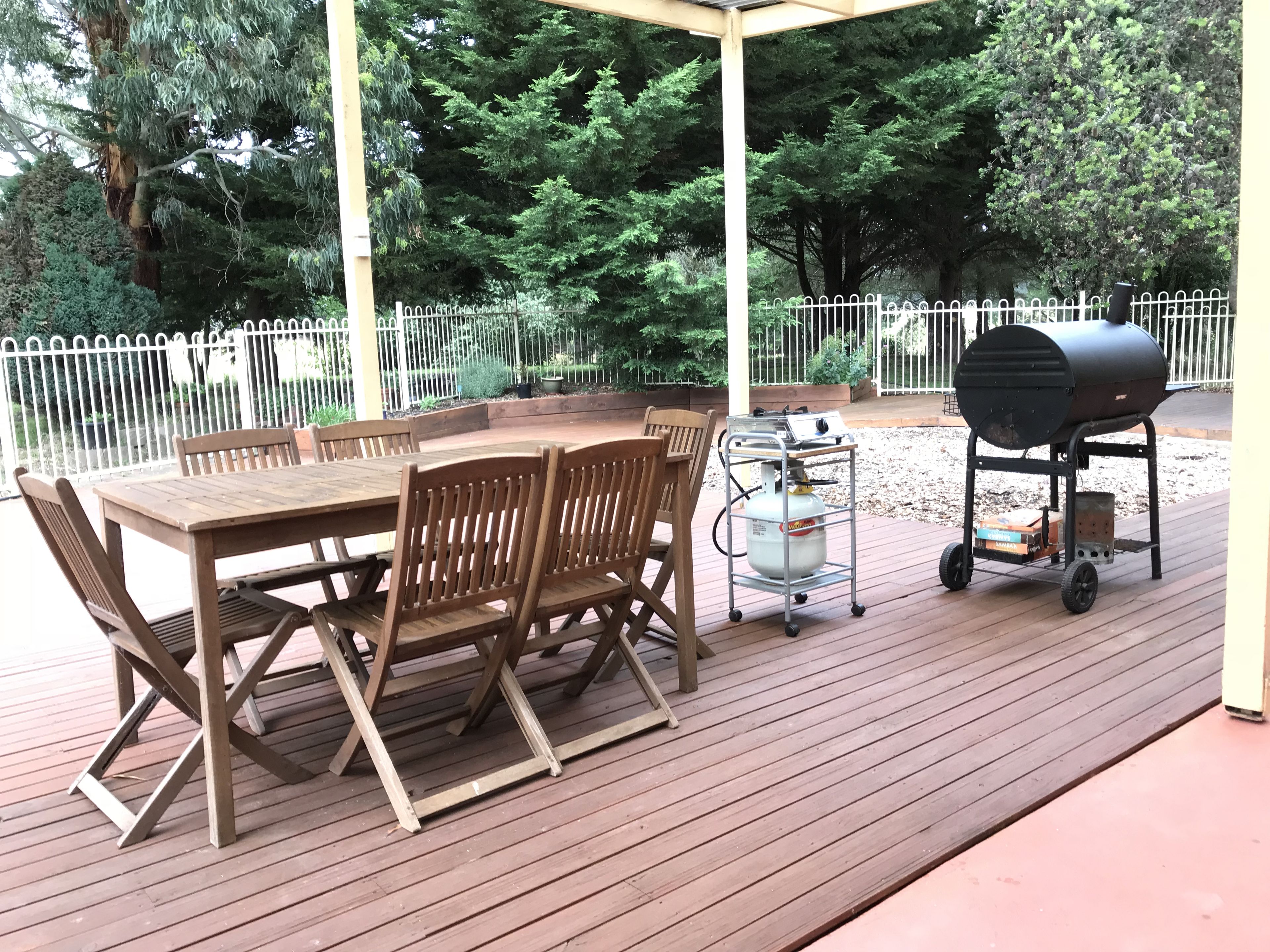 Um pátio externo com mesa e cadeiras de madeira, churrasqueira e um cilindro de gás em um carrinho.