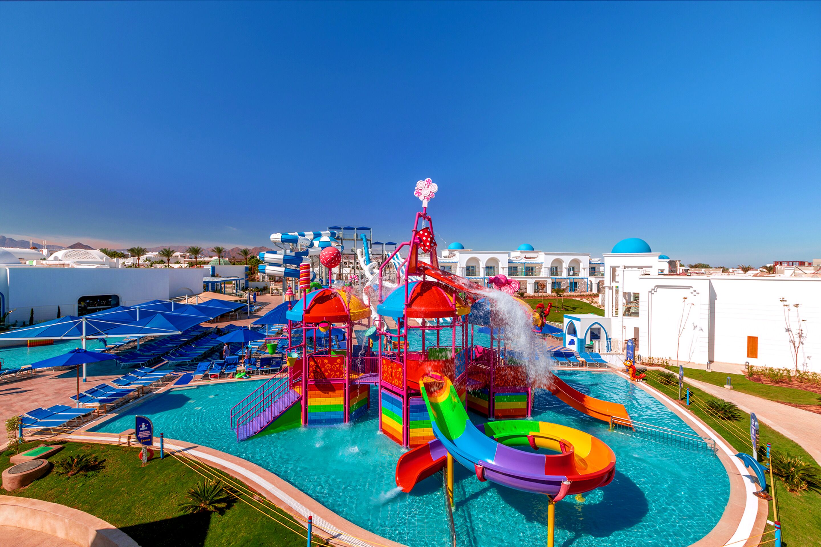 Um parque aquático com vários toboáguas e piscinas, cercado por edifícios brancos com cúpulas azuis.