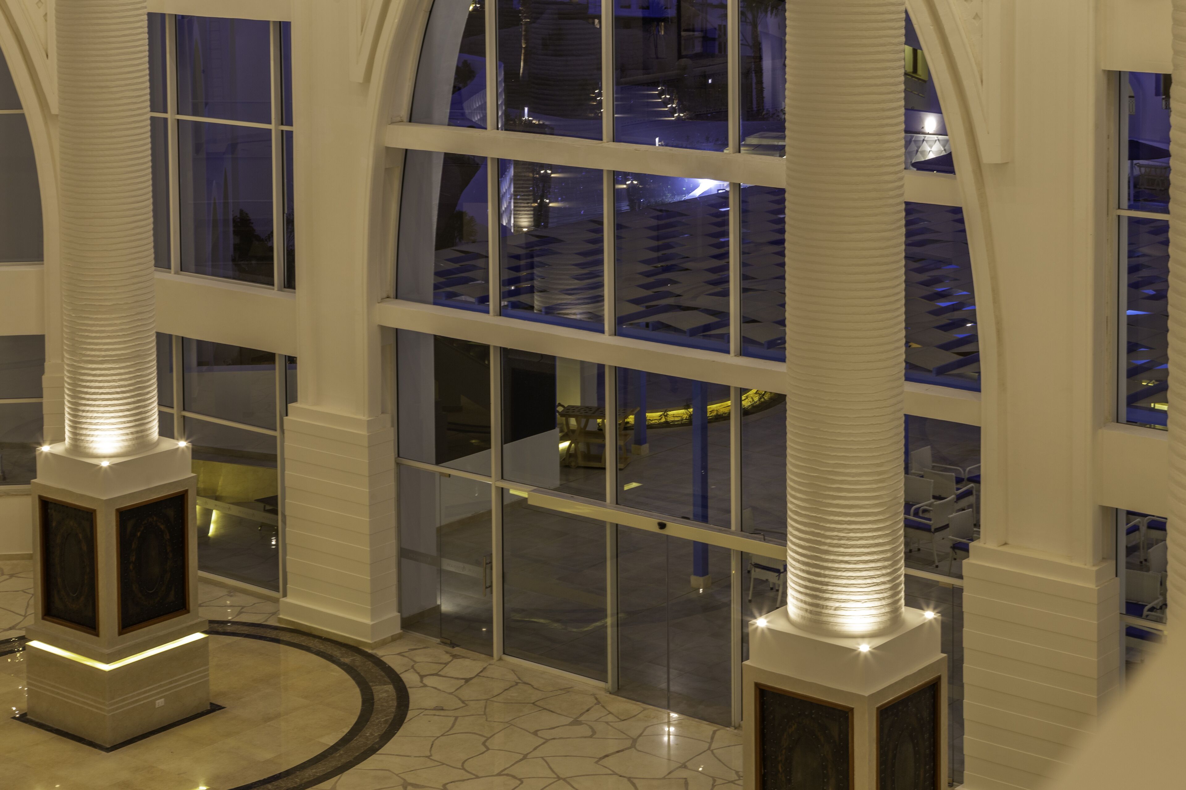 Um hall espaçoso com amplas janelas, piso de mármore circular e duas colunas altas com base iluminada.