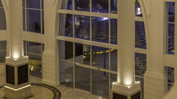 Um hall espaçoso com amplas janelas, piso de mármore circular e duas colunas altas com base iluminada.