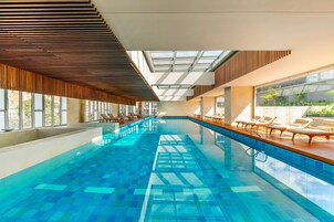 Piscina interior moderna com azulejos azuis translúcidos, deck de madeira e espreguiçadeiras.