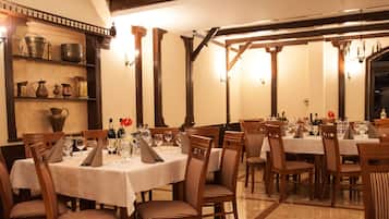 Uma sala de jantar com várias mesas preparadas para hóspedes, cadeiras de madeira e uma estante com objetos decorativos.