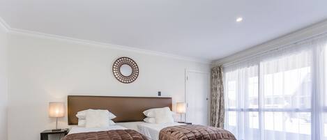Quarto de hotel com duas camas, uma janela grande com cortinas leves, um espelho decorativo na parede e piso coberto de carpete.