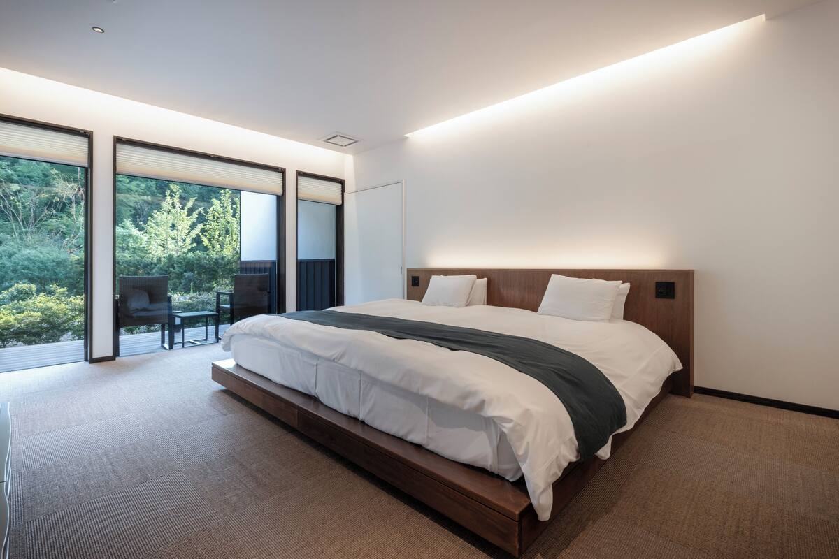 Quarto moderno com uma cama grande, cabeceira de madeira e vista para o verde através de amplas janelas.