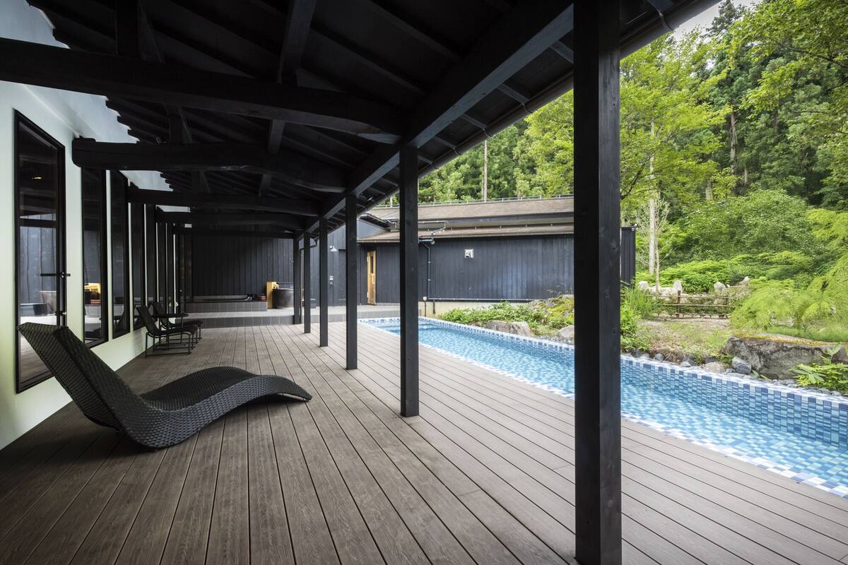 Área externa coberta com piscina, deck de madeira, espreguiçadeiras e vista para uma área arborizada.