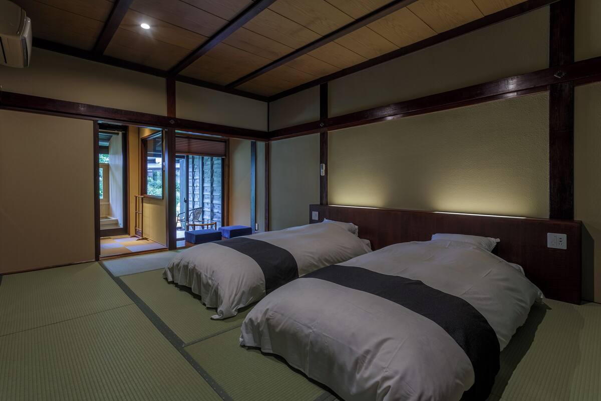 Um quarto japonês tradicional com tatames, duas camas com roupa de cama escura e clara, teto de madeira e uma porta deslizante que dá para uma área externa.