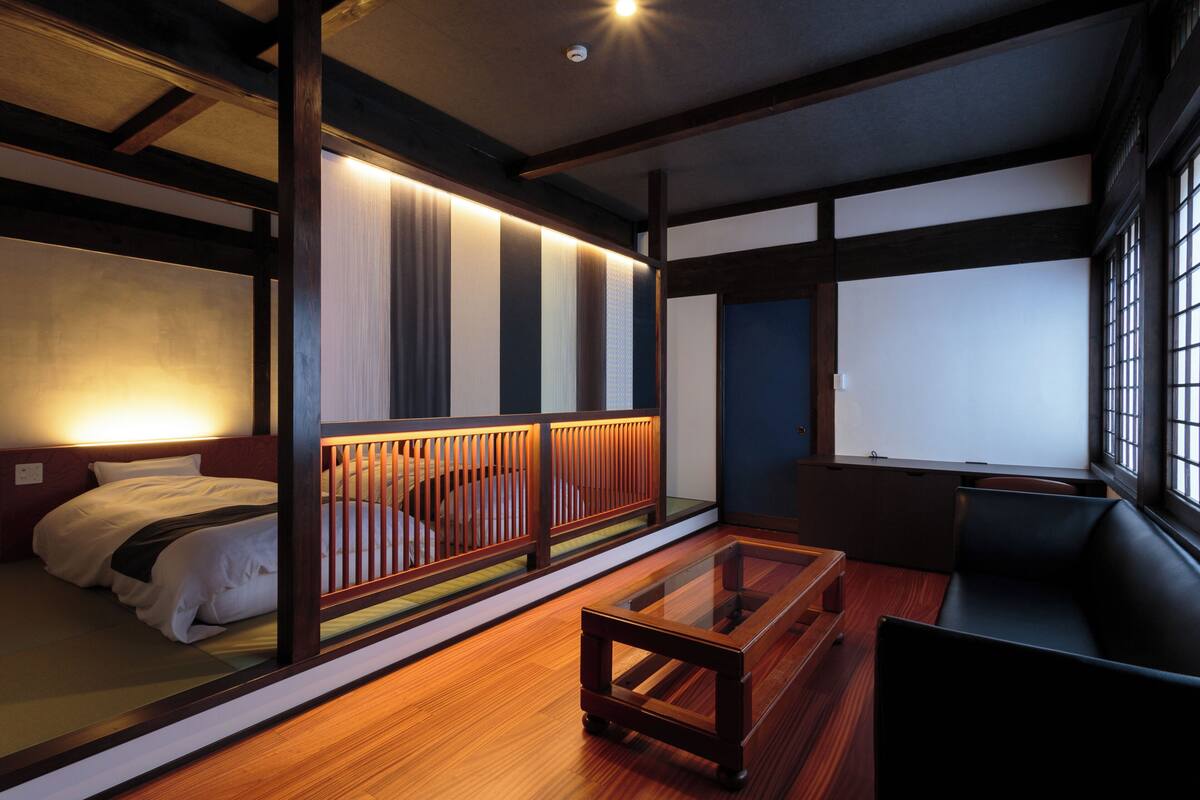 Um quarto tradicional japonês com piso de tatame, uma mesa de madeira baixa, um sofá e uma porta deslizante com vista para o exterior.