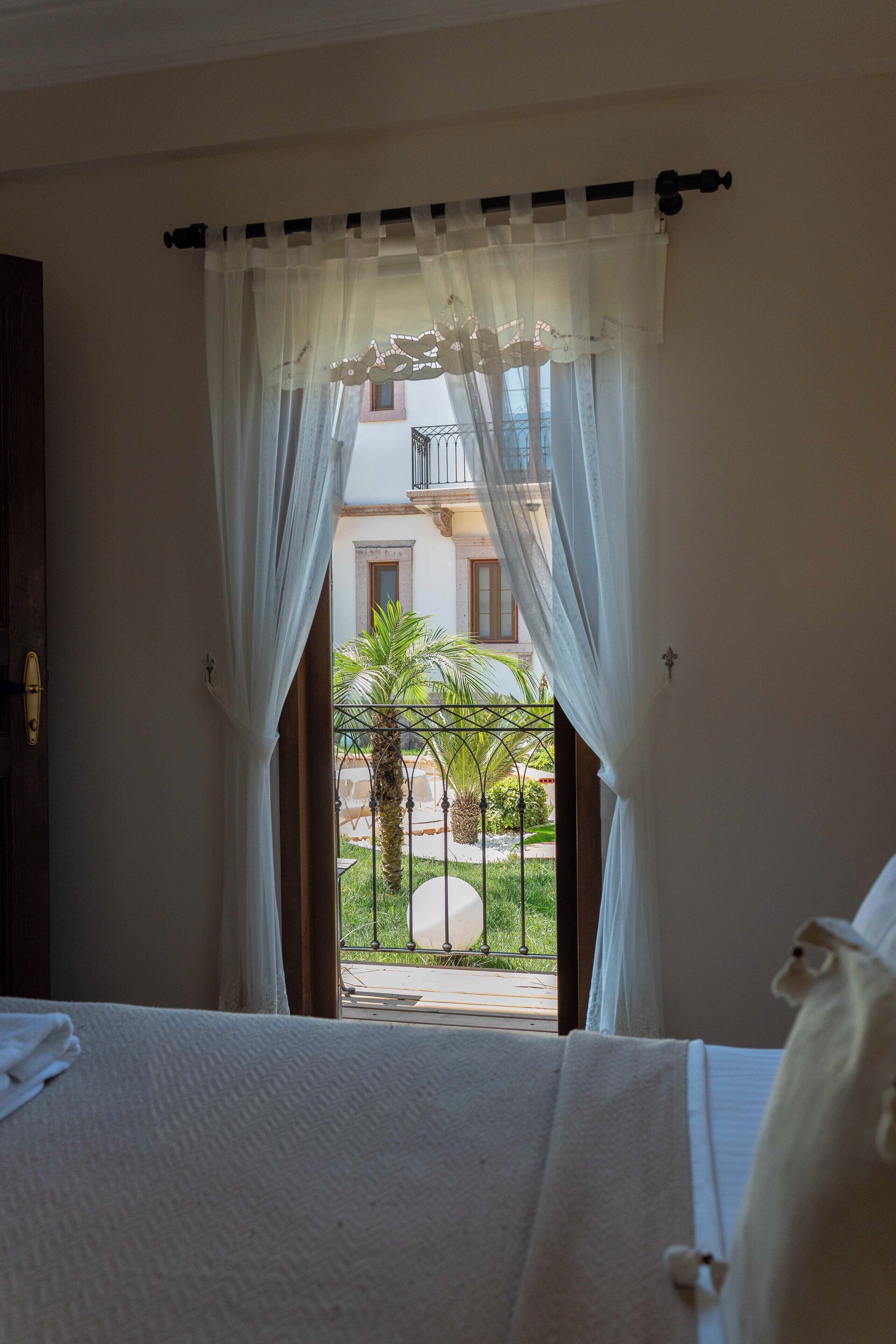 Quarto com uma cama, cortinas brancas e vista para uma varanda com palmeira e um edifício ao fundo.