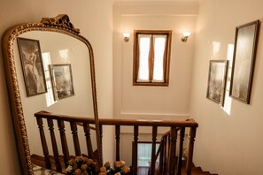 Uma escadaria com corrimão de madeira e um espelho grande e ornamentado. Quadros emoldurados nas paredes e uma janela com cortinas.