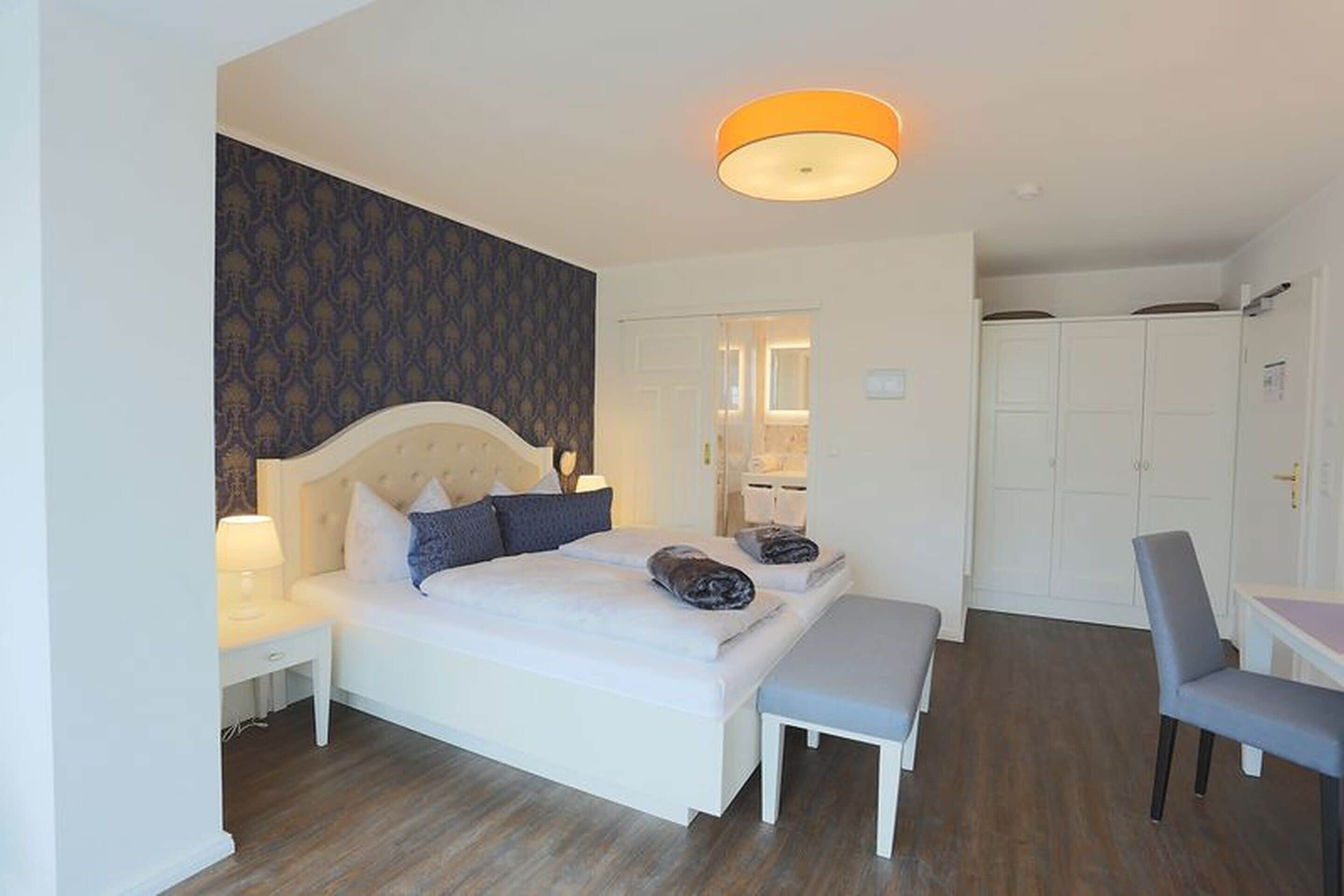 Quarto de hotel com uma cama grande, uma mesa de cabeceira com abajur, um banco, uma cadeira e um guarda-roupa.