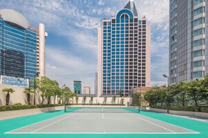 Uma quadra de tênis ao ar livre rodeada por edifícios modernos, com céu claro e alguma vegetação.