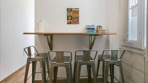 Quarto pequeno com uma mesa de madeira e quatro cadeiras de metal. Há um cartaz na parede e uma janela à direita.
