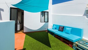 Uma área de estar moderna ao ar livre, com um sofá azul e almofadas, uma mesa pequena e piso de grama sintética verde.
