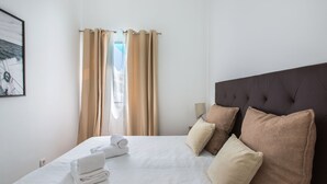 Cama bem arrumada com lençóis brancos e toalhas dobradas, cabeceira escura e almofadas bege.
