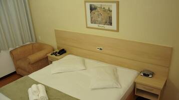 Quarto de hotel com uma cama, uma cadeira, mesa de cabeceira com telefone e um quadro na parede.