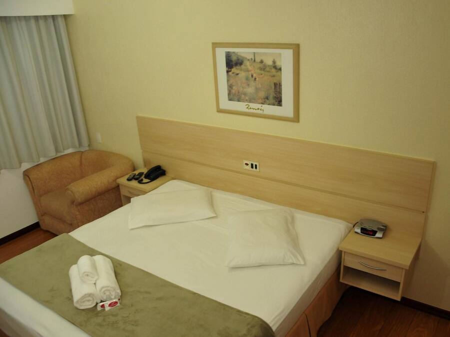 Quarto de hotel com uma cama, uma cadeira, mesa de cabeceira com telefone e um quadro na parede.