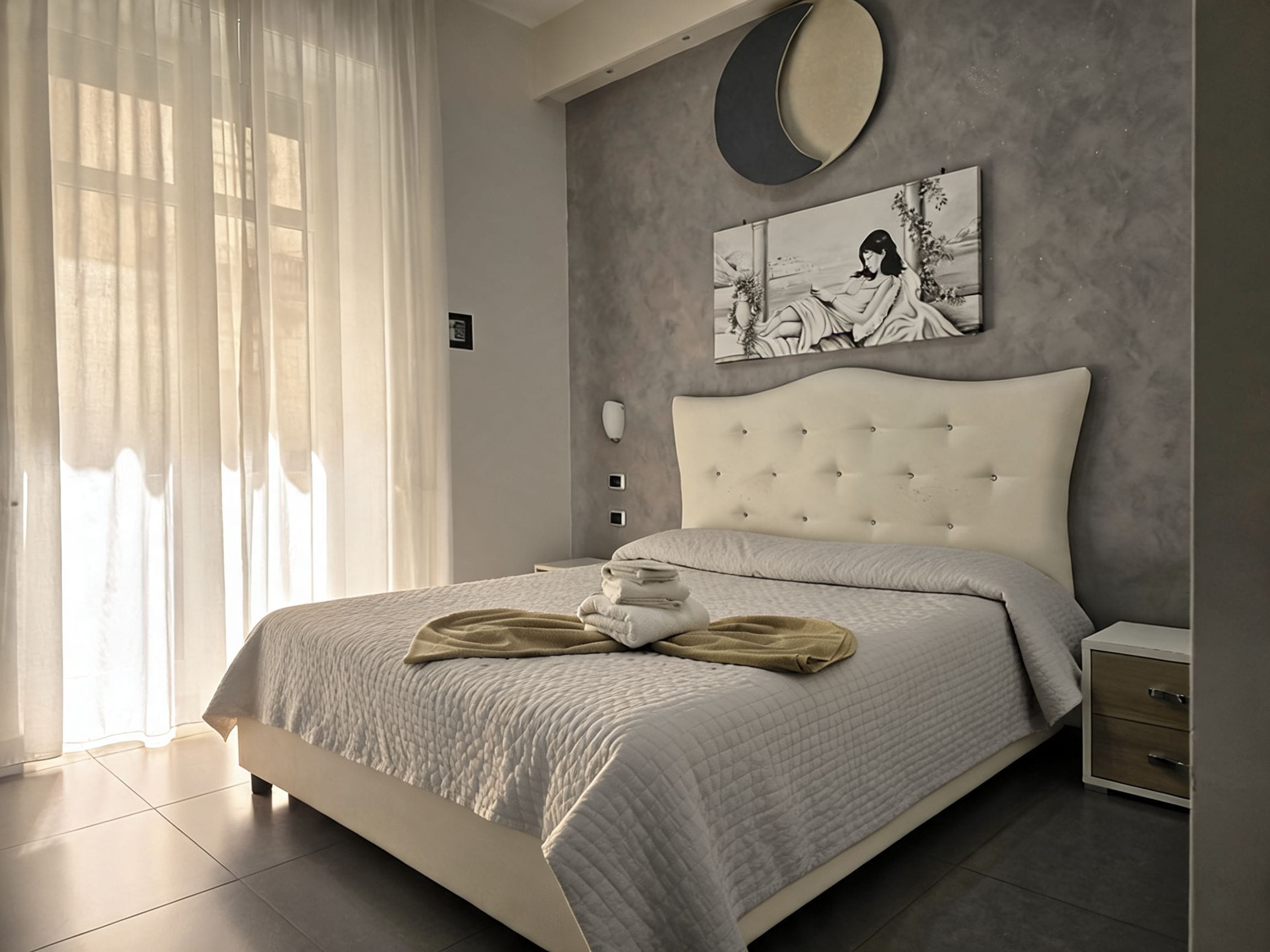 Quarto com cama, mesa de cabeceira, relógio de parede e janela com cortinas.