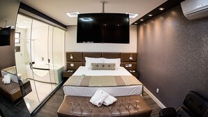 Quarto de hotel moderno com uma cama grande, TV de tela plana, chuveiro em box de vidro e um espaço de estar com um banquinho de couro marrom.