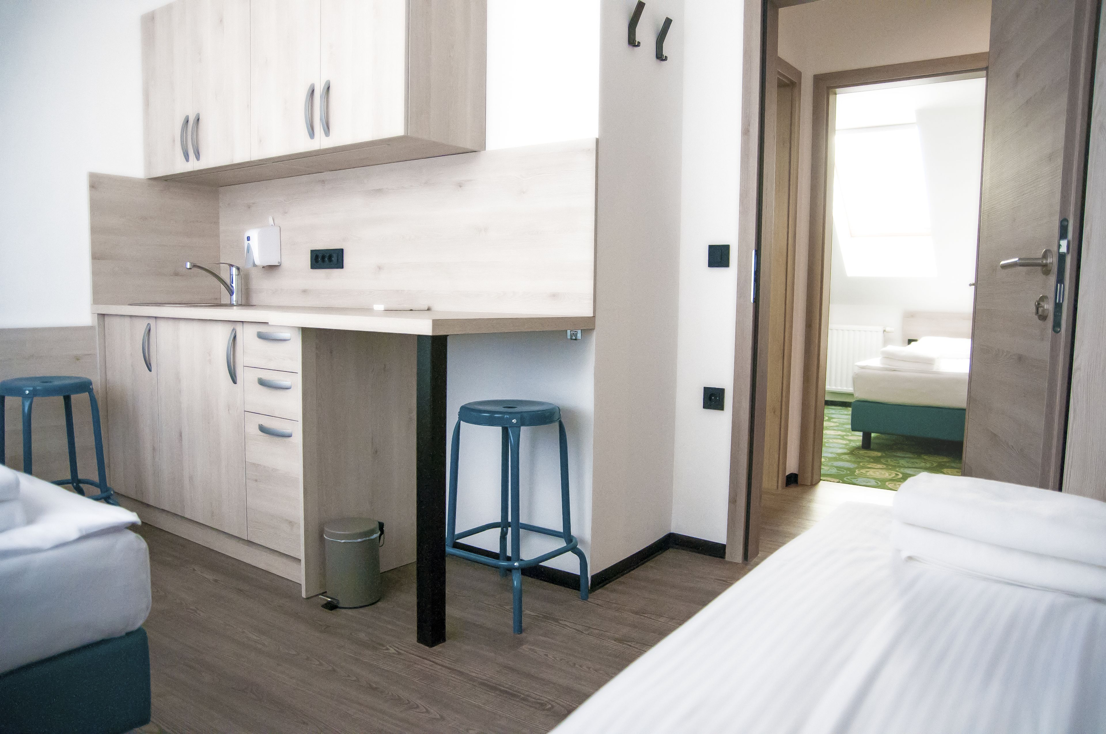 Quarto de hotel pequeno com cozinha compacta, uma cama, um banquinho e vista para um quarto com carpete verde.