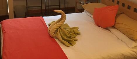 Cama bem arrumada com colcha vermelha e bege, toalha em formato de cisne e dois travesseiros laranja.