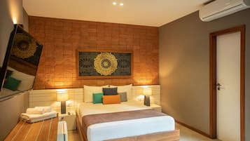 Quarto de hotel moderno com parede de tijolos, uma cama grande com várias almofadas, luminárias laterais, uma mesinha de madeira com um telefone e uma porta.