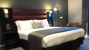 Quarto de hotel com uma cama grande, cabeceira escura, luminárias laterais e mesa de cabeceira.