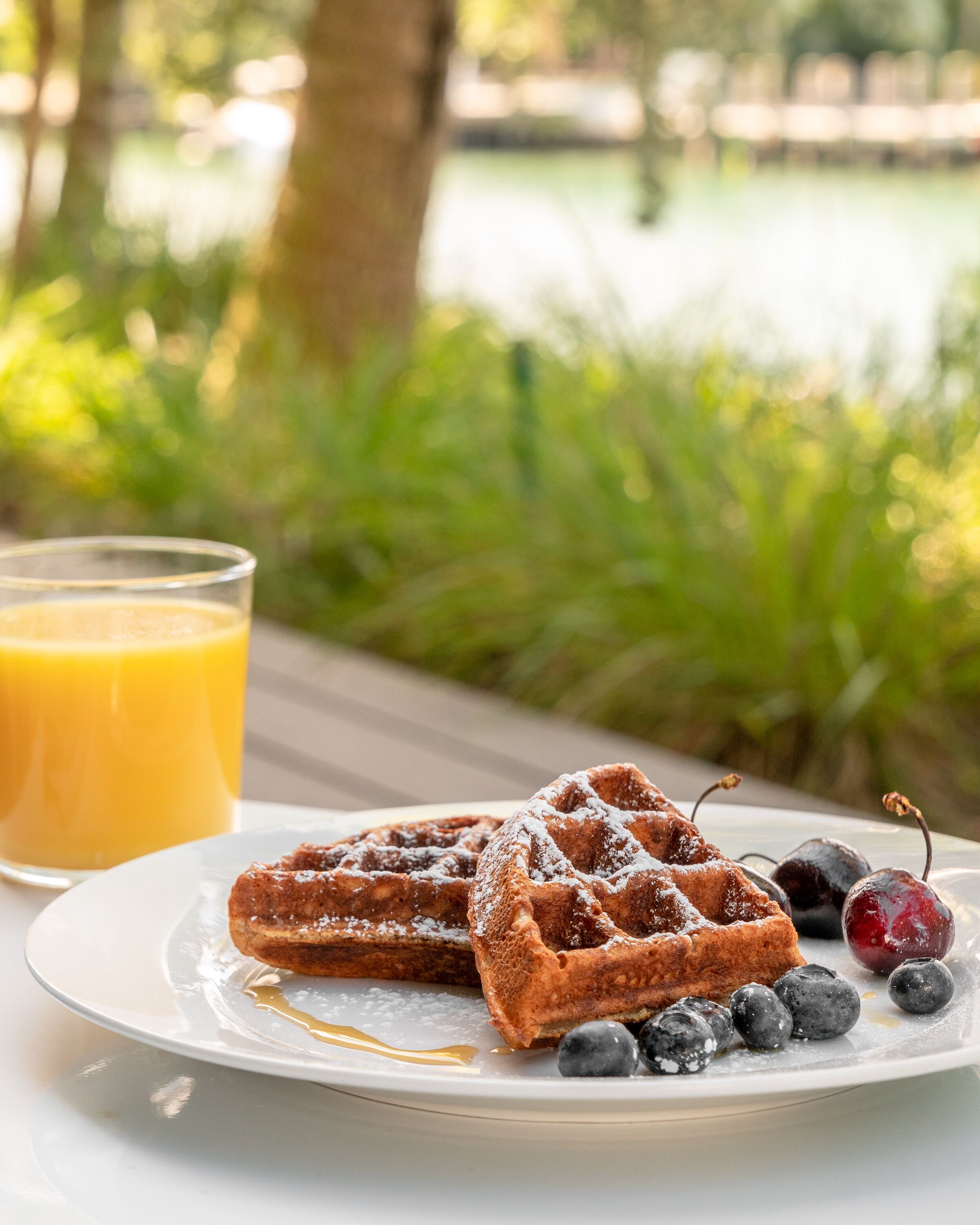 Waffles com açúcar em pó e xarope, mirtilos e um copo de sumo de laranja numa placa branca.